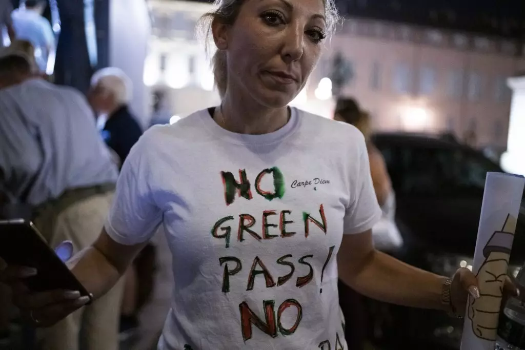 Mae arddangoswr yn dal Tocyn Gwyrdd ar grys-t yn ystod y Protest No Vax ar Orffennaf 22, 2021 yn Turin, yr Eidal. Protest gan No Vax neu Vax Am Ddim yn erbyn cyflwyno tocyn iechyd, a elwir yn docyn gwyrdd gan Lywodraeth yr Eidal a fydd yn orfodol i gael mynediad i byllau nofio, campfeydd a neuaddau chwaraeon, digwyddiadau chwaraeon, cyngherddau, ffeiriau a lleoliadau diwylliannol gan gynnwys amgueddfeydd, sinemâu , a theatrau. Llun gan Stefano Guidi, Getty Images News.