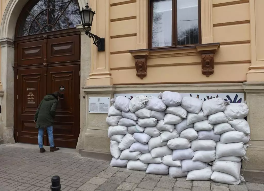 Gizon bat zutik dagoen Lviveko Arte Galeria Nazionaleko ate batetik begiratzen du, hare-poltsez babestuta dauden leihoak dituena, 2022ko martxoaren 7an, Ukrainako mendebaldeko Lviv hirian, Errusiak Ukrainaren aurkako inbasio militarra hasi eta 12 egunera. Argazkia: Yuriy Dyachyshyn / AFP Getty Images bidez.