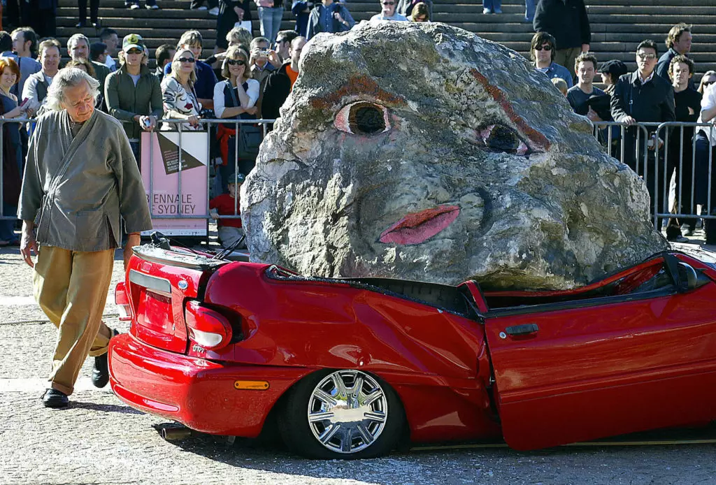 L'artiste américain Jimmie Durham (L), inspecte une voiture qui a été écrasée avec un rocher de granit de six tonnes peint avec un visage humain devant une grande foule à l'Opéra de Sydney, le 05 juin 2004, comme une œuvre qui fait partie de la Biennale de Sydney. La Biennale, le premier événement international d'arts visuels contemporains d'Australie, se tiendra du 04 juin au 15 août et présentera le travail de cinquante et un artistes de trente-deux pays, avec des expositions présentées autour de l'estran de Sydney, des galeries d'art et des musées. AFP PHOTO/Greg WOOD (Le crédit photo doit se lire GREG WOOD/AFP/Getty Images)