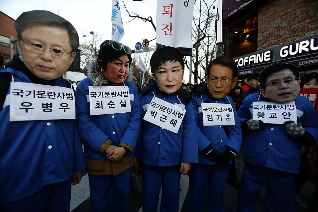 Ndị na-eme ngagharị iwe na Seoul na Disemba 10, 2016 na-eyi mkpuchi ihu Choi Soon-Sil (n'etiti aka ekpe) na Park Geun-Hye (n'etiti aka nri). Ka onye isi ala na-eche ikpe maka nchụpụ, ndị omebe iwu na-amalite inyocha aha onye omenkà eboro ebubo nke ndị omenaala 9,000. Foto sitere n'ikike Chung Sung-Jun/Getty Images.