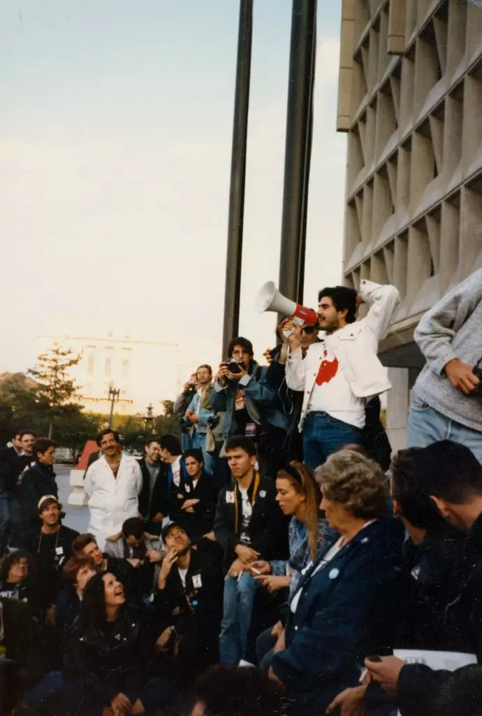 Gregg Bordowitz com um megafone dirigindo-se a uma multidão de manifestantes em frente à Food and Drug Administration em 1988. Várias pessoas estão rindo na foto, e um senso de humor – às vezes distorcido – paira sobre sua retrospectiva do MoMA PS1. Cortesia do artista.