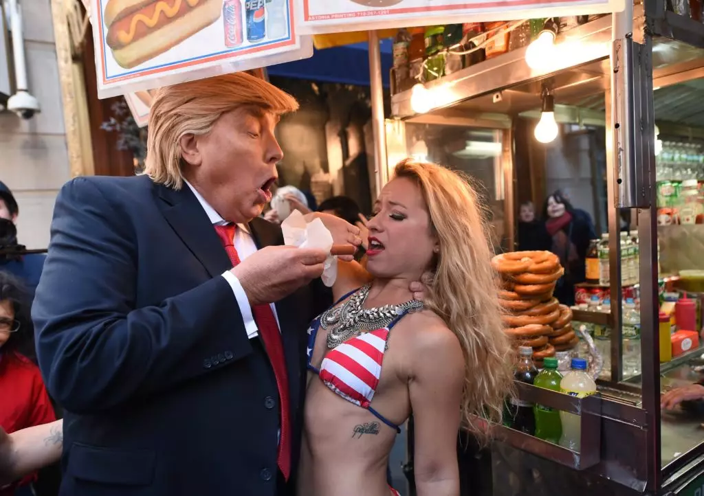 Alison Jekson ijrosidagi sanʼat asarida Donald Tramp rolini oʻynagan aktyor boshqa bir ijrochini hot-dog bilan oziqlantirmoqda. Timoti A. Klari/AFP/Getty Imagesning izni bilan.