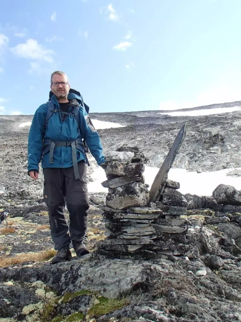 Seorang arkeolog dengan salah satu tugu batu yang menandai celah gunung di Lendbreen. Batuan berwarna terang di latar belakang tertutup salju dan es hingga saat ini. Foto oleh James Barrett, Universitas Cambridge.