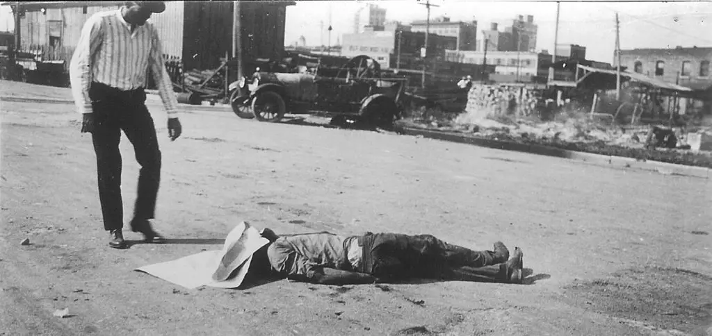 Kroppen av ett oidentifierat svart offer för massakern i Tulsa Race ligger på gatan när en vit man står över honom, Tulsa, Oklahoma, 1 juni 1921. Foto av Greenwood Cultural Center/Getty Images.