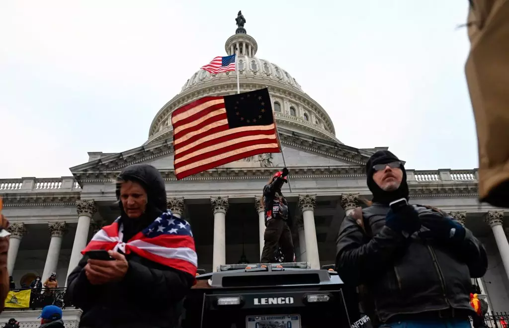 Yon patizan Trump balanse yon drapo pandan li kanpe sou yon machin gouvènman an devan Kapitòl Ameriken an nan Washington DC nan dat 6 janvye 2021. Manifestan yo te kraze sekirite epi yo te antre nan kapital la pandan Kongrè a t ap debat sou Sètifikasyon Vòt Elektoral pou eleksyon prezidansyèl 2020 la. (Foto pa Andrew Caballero-Reynolds/AFP/Getty Images.