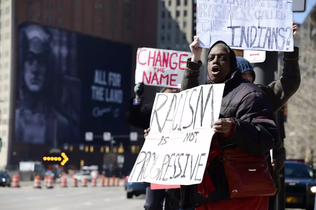 Aktivisten protestieren gegen die Verwendung des Cleveland Indians-Maskottchens Chef Wahoo vor dem Spiel zwischen den Cleveland Indians und den Chicago White Sox im Progressive Field während des Indians Home Opener am 1. April 2019 in Cleveland, Ohio. Im Juli 2021 gab das Team bekannt, dass es seinen Namen 2022 in Guardians ändern werde. Im Hintergrund ist ein öffentliches Kunstbanner mit einem der Guardians of Traffic, dem neuen Namensvetter des Teams, zu sehen. Foto von Jason Miller/Getty Images.